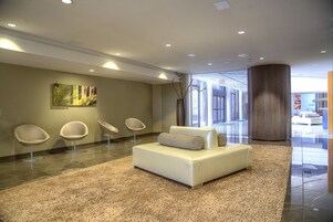 Um lobby moderno com um sofá branco, três cadeiras brancas, um vaso grande e um tapete texturizado.