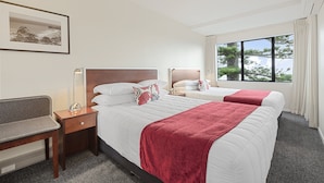 Quarto de hotel com duas camas, uma escrivaninha, uma cadeira, mesa de cabeceira, abajur e janela com vista para árvores.