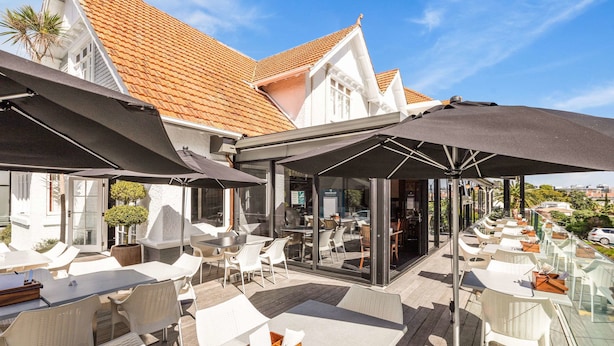 Um pátio externo com móveis de vime branco, guarda-sóis pretos e um banco de madeira.