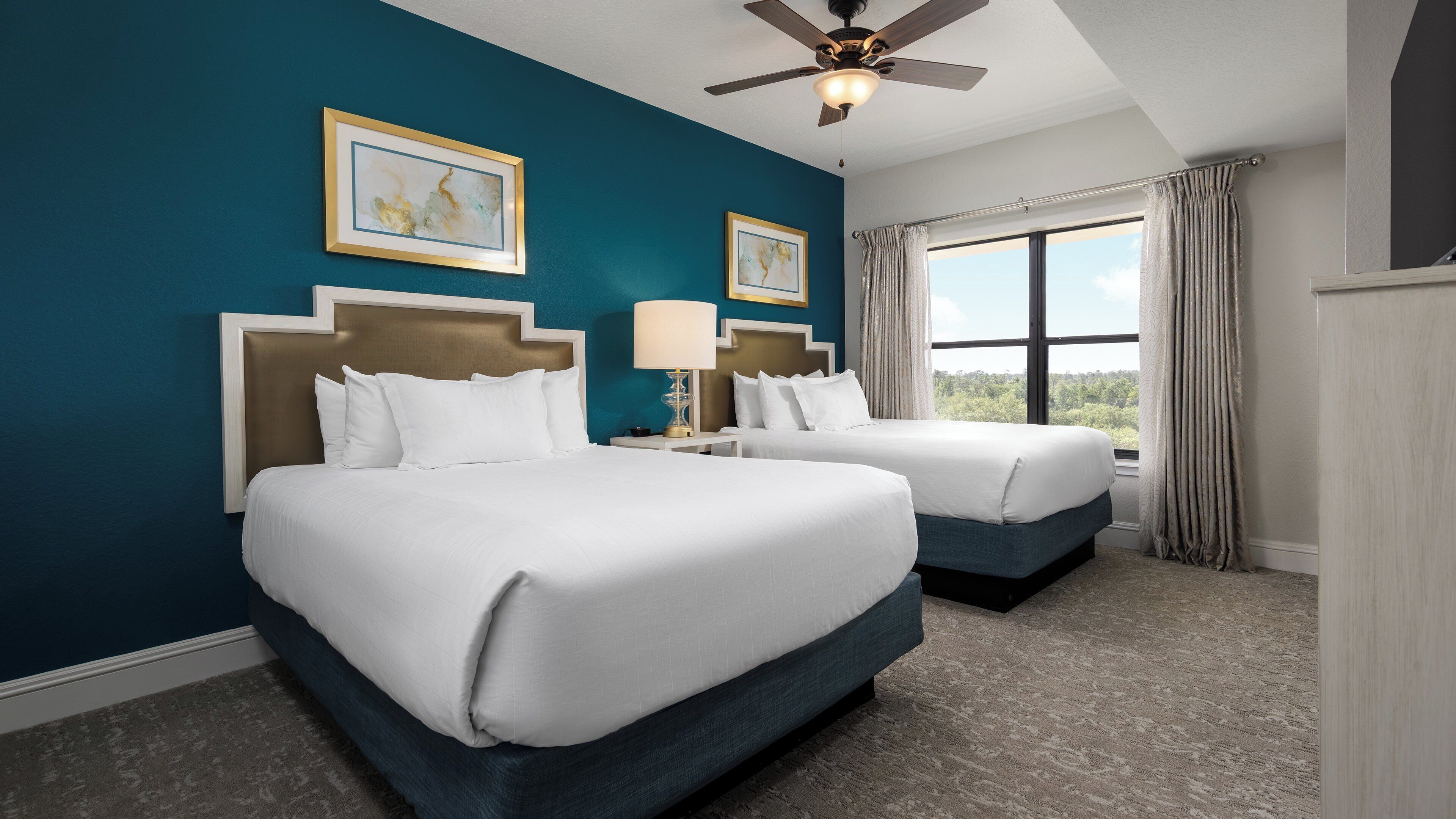 Quarto de hotel com duas camas, ventoinha no teto, grande janela com cortinas e obras de arte nas paredes.