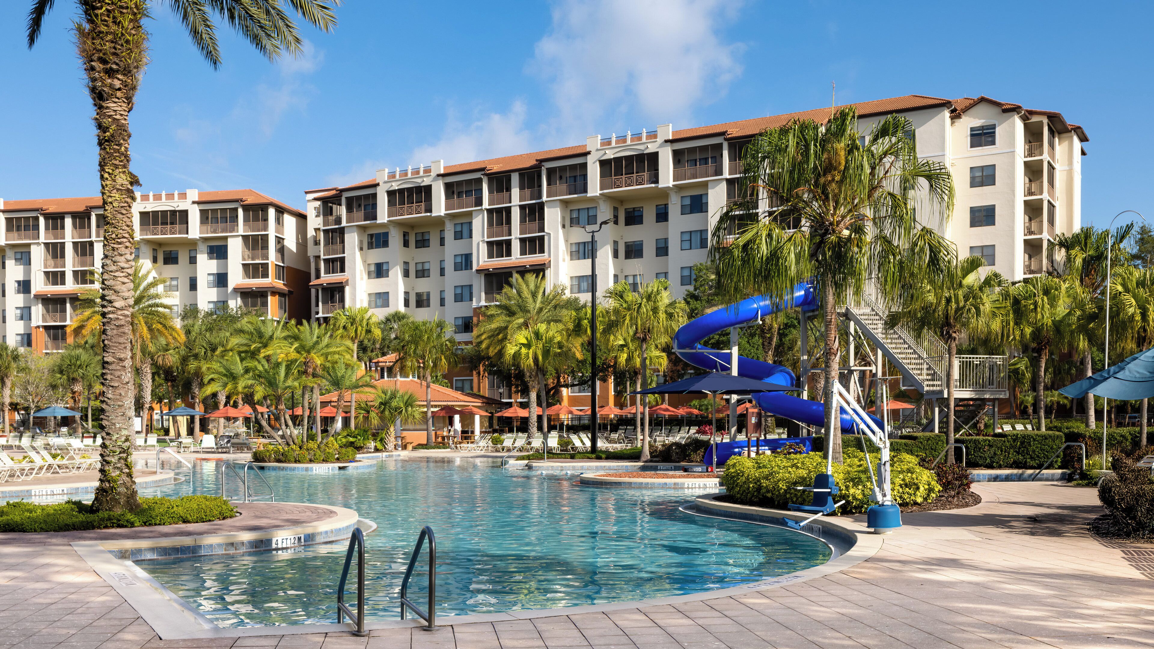 Um hotel com piscina, palmeiras e um escorrega aquático azul.