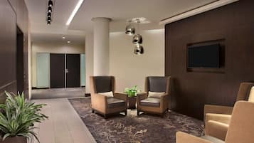 Um hall de entrada com duas poltronas, uma mesinha com uma planta, um tapete estampado e uma televisão na parede.
