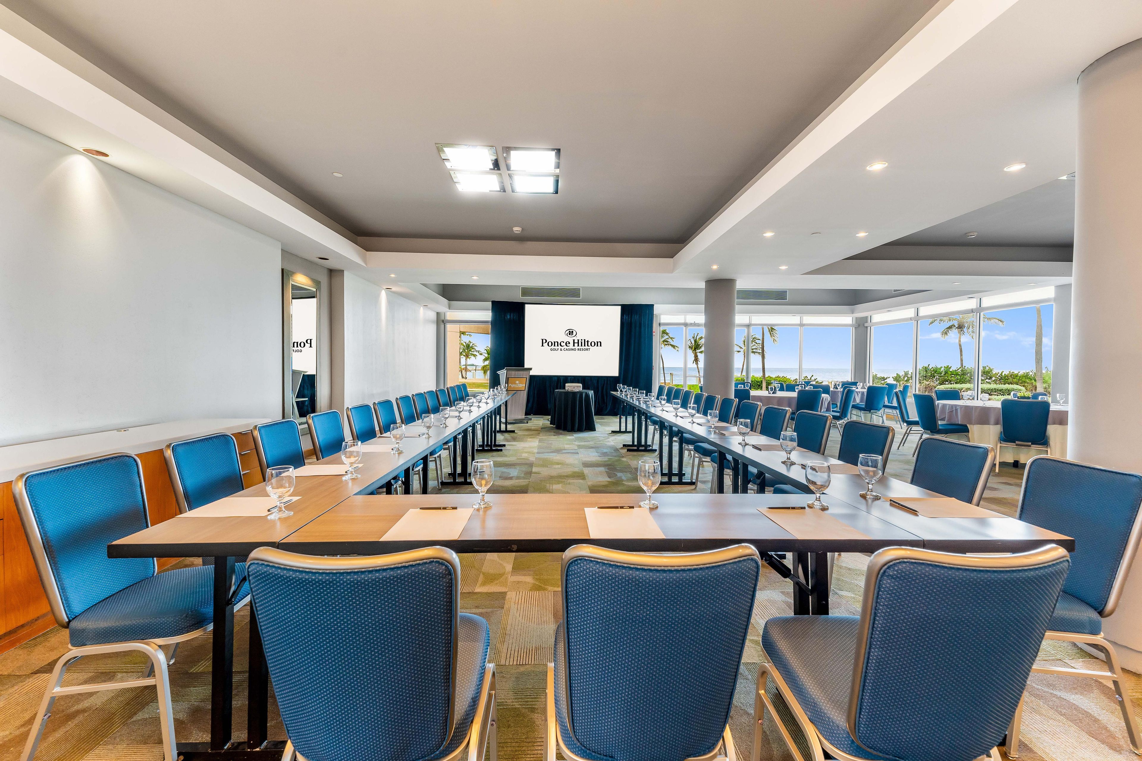 Uma sala de conferências com uma mesa comprida, cadeiras azuis e uma tela de apresentação na frente.