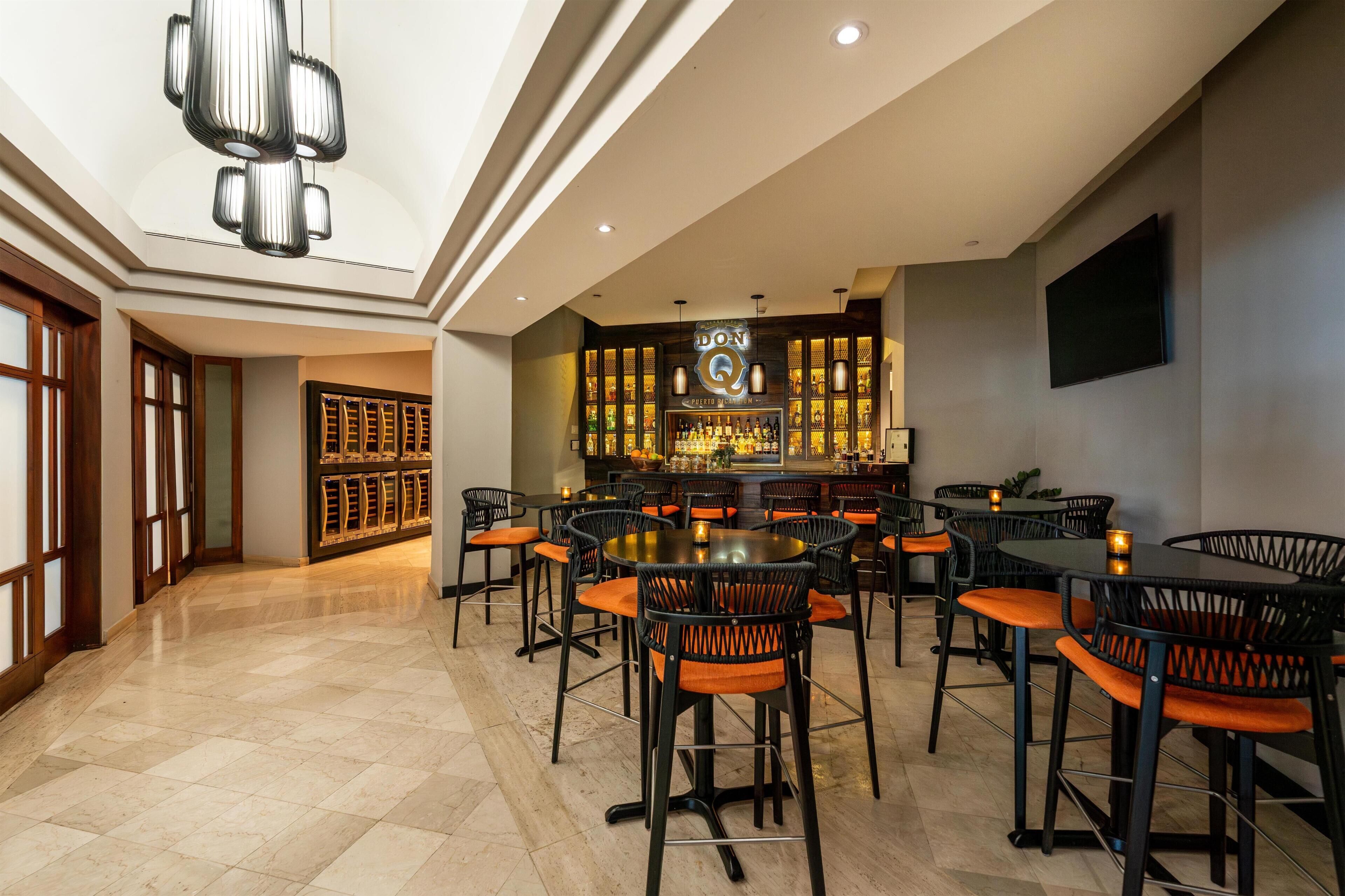 Uma área de bar ampla, com mesas e cadeiras redondas, balcão e um grande lustre.