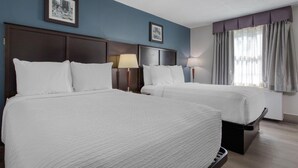 Quarto de hotel com duas camas, cabeceira, mesa de cabeceira com abajur, janela com cortinas e duas imagens emolduradas na parede.