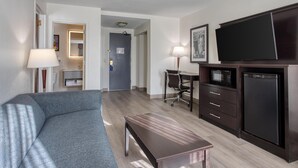 Quarto de hotel com sofá cinzento, mesa de centro de madeira, escrivaninha com cadeira, televisão de tela plana, micro-ondas e geladeira.