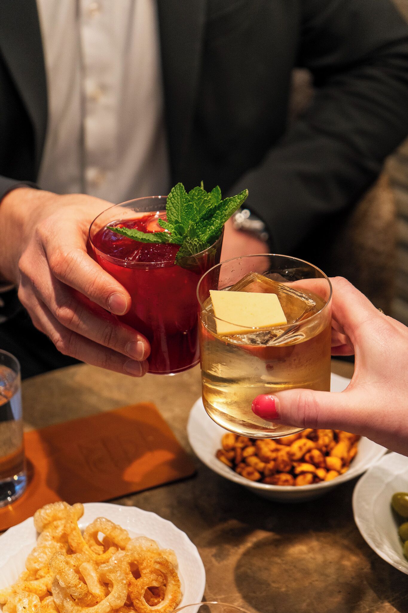 Duas pessoas segurando copos com bebidas, uma com um drink vermelho e um ramo de hortelã, a outra com um drink transparente e um cubo de gelo.