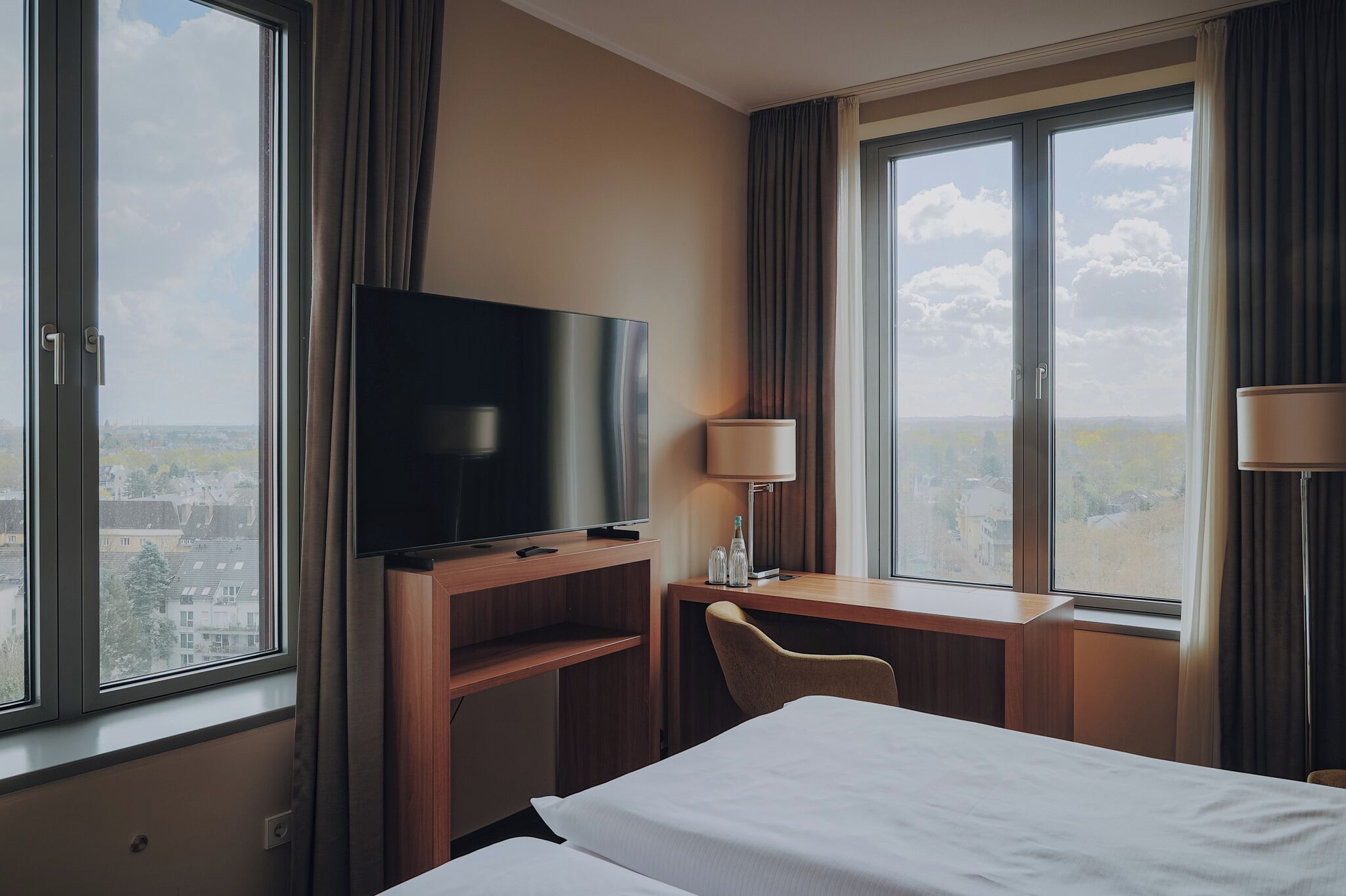 Quarto de hotel com uma cama, uma televisão sobre uma escrivaninha de madeira, uma cadeira e janelas amplas com cortinas.