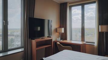 Quarto de hotel com uma cama, uma televisão sobre uma escrivaninha de madeira, uma cadeira e janelas amplas com cortinas.