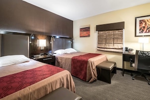 Quarto de hotel com duas camas, uma escrivaninha, uma cadeira, janela com persianas e um quadro na parede.