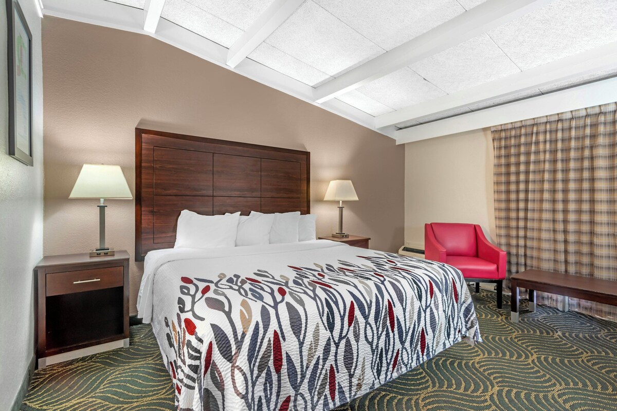 Quarto de hotel com uma cama grande, duas mesas de cabeceira com luminárias, uma cadeira vermelha, um banco e um tapete estampado.