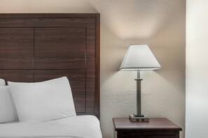 Uma cama com roupa de cama branca, cabeceira de madeira, mesa de cabeceira com luminária e parede com textura.
