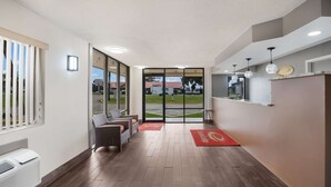 Uma área de recepção com piso de madeira, balcão de atendimento, espaço de estar com cadeiras e vista para o exterior pelas portas de vidro.