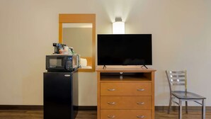 Quarto compacto com televisão de tela plana sobre um móvel de madeira, micro-ondas sobre um suporte preto pequeno, uma cadeira e um espelho com luz acima.