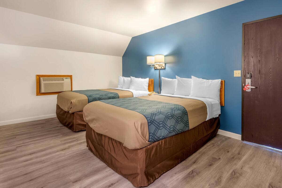 Quarto de hotel com duas camas, uma parede azul decorativa, uma porta de madeira e um ar condicionado fixado na parede.