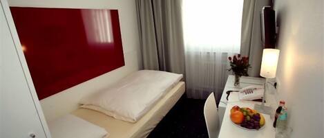 Quarto de hotel com uma cama de solteiro, uma escrivaninha com luminária, uma cadeira, uma mesa com frutas e um jornal, e uma janela com cortinas.