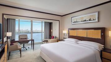 Quarto de hotel com uma cama grande, uma escrivaninha, uma cadeira, uma televisão e vista da cidade por grandes janelas.