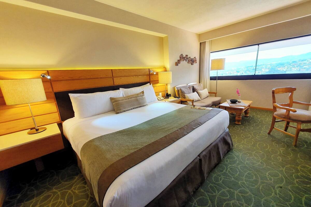 Quarto de hotel com uma cama grande, uma escrivaninha, uma cadeira e vista para a cidade pela janela.