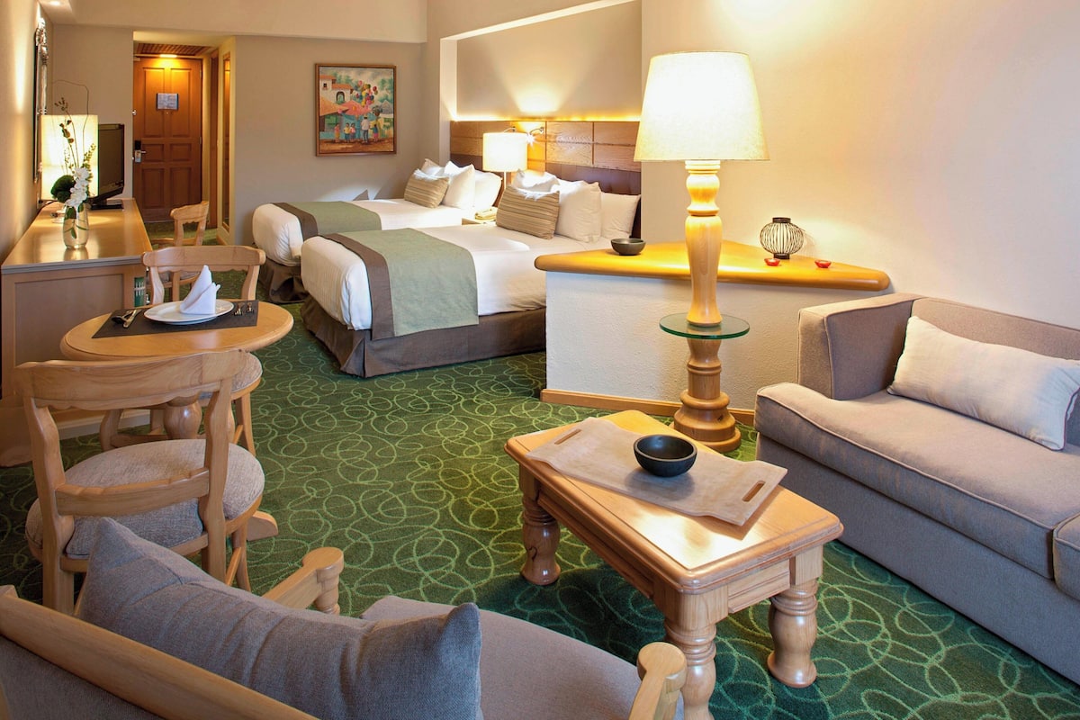 Quarto de hotel com duas camas, um espaço de estar com sofá e poltronas, uma mesinha com abajur, e uma escrivaninha com telefone e um vaso de flores.