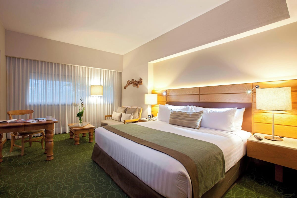 Quarto de hotel com uma cama grande, uma escrivaninha, uma cadeira, um abajur e uma janela com cortinas.