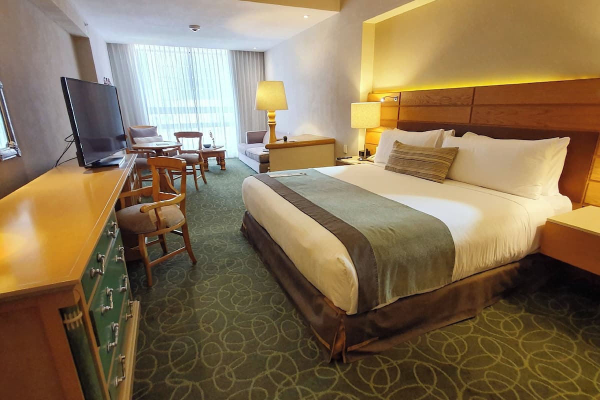 Quarto de hotel com cama, escrivaninha, televisão, cadeiras e vista para o exterior.