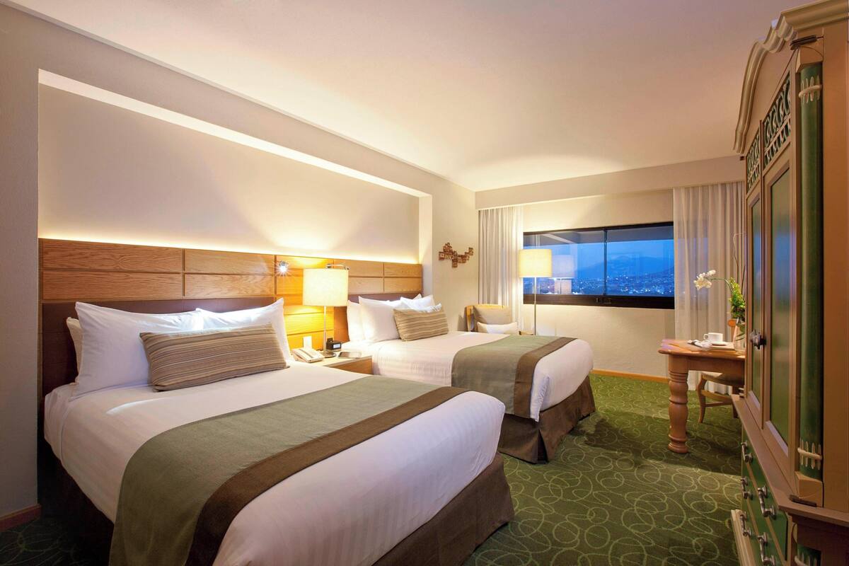 Quarto de hotel com duas camas, mesa de cabeceira, escrivaninha e vista para a cidade.