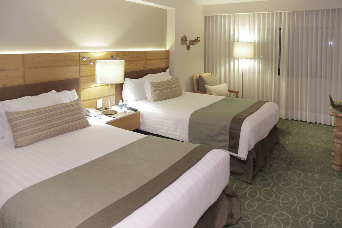 Quarto de hotel com duas camas, uma mesa de cabeceira com luminária, uma cadeira e uma janela grande com cortinas.