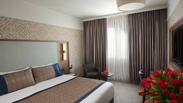 Quarto de hotel com cama grande, escrivaninha, cadeira, mesa com frutas e um vaso de flores.