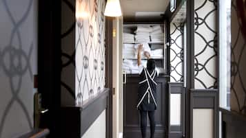 Uma pessoa vestida de preto e branco alcança toalhas em um corredor com paredes estampadas e uma porta que dá para um quarto com cama.