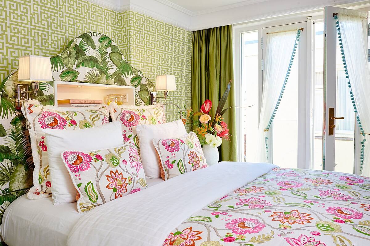 Quarto de hotel com cama, almofadas estampadas com motivos florais, mesa de cabeceira com luminárias e vista para o exterior.