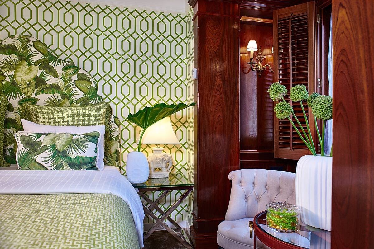 Quarto de hotel com papel de parede verde e branco estampado, uma cama com roupa de cama verde e branca, uma poltrona branca, uma mesinha com abajur e uma planta grande.