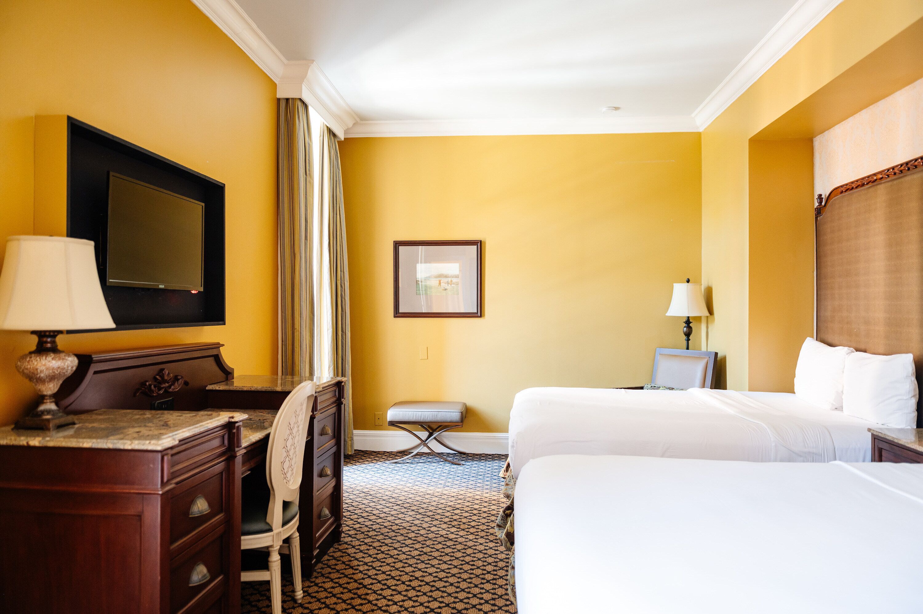 Quarto de hotel com duas camas, uma escrivaninha com luminária, uma cadeira, uma televisão e um quadro pendurado na parede.