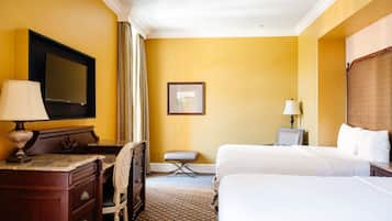 Quarto de hotel com duas camas, uma escrivaninha com luminária, uma cadeira, uma televisão e um quadro pendurado na parede.