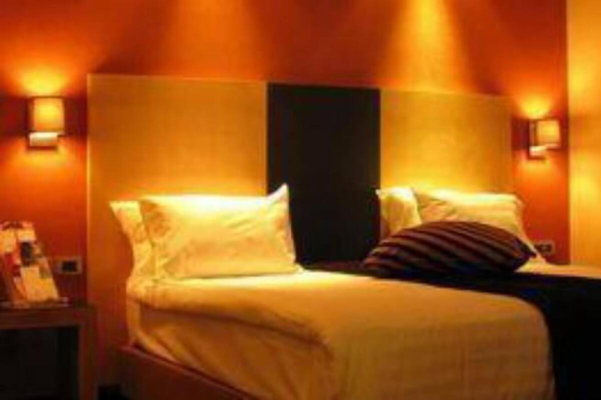 Quarto de hotel com cama, almofadas, mesa de cabeceira e luminárias fixadas na parede.
