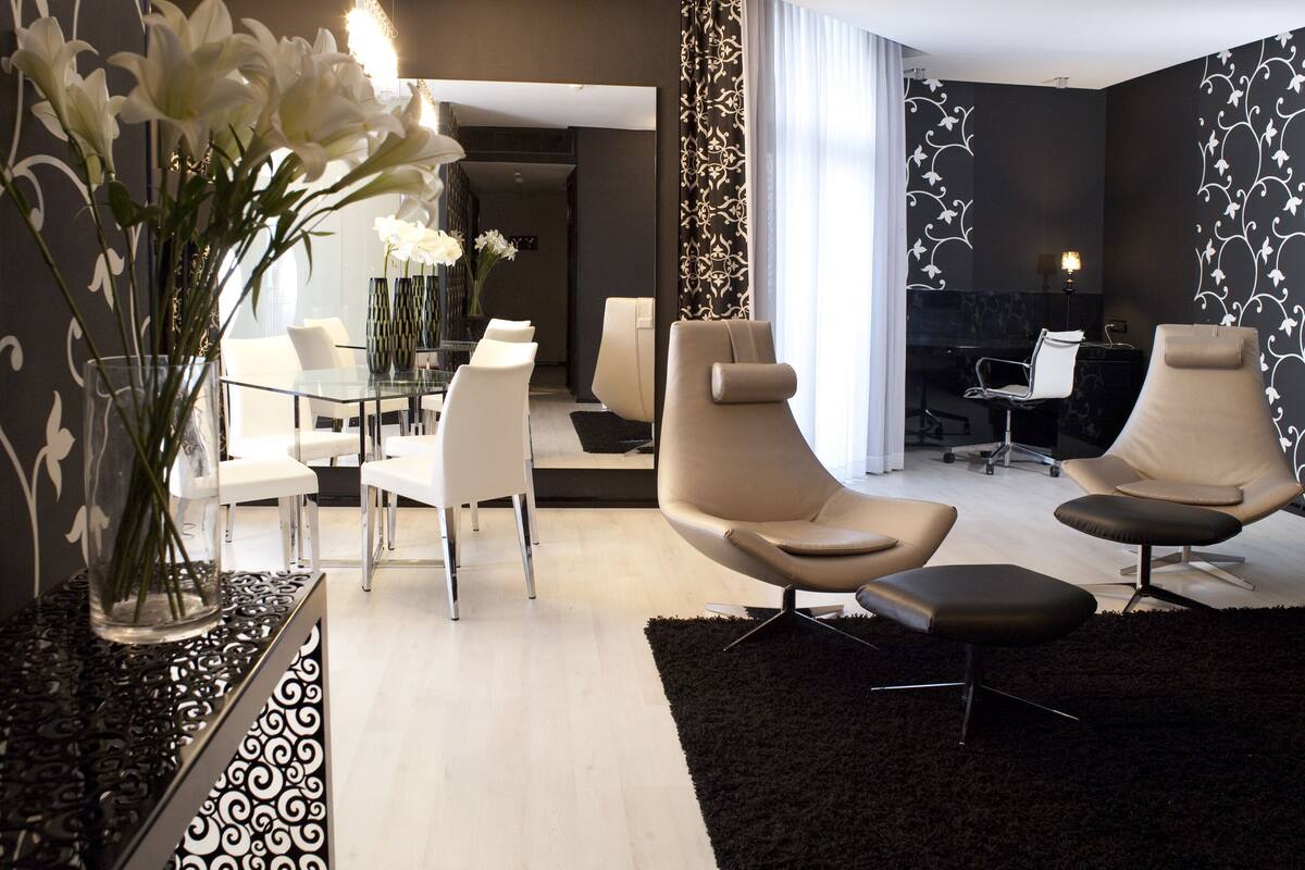Uma sala de estar moderna com uma mesa de centro de vidro, cadeiras brancas e um tapete preto. Há duas poltronas bege e um sofá. Um vaso com flores brancas está sobre um console.