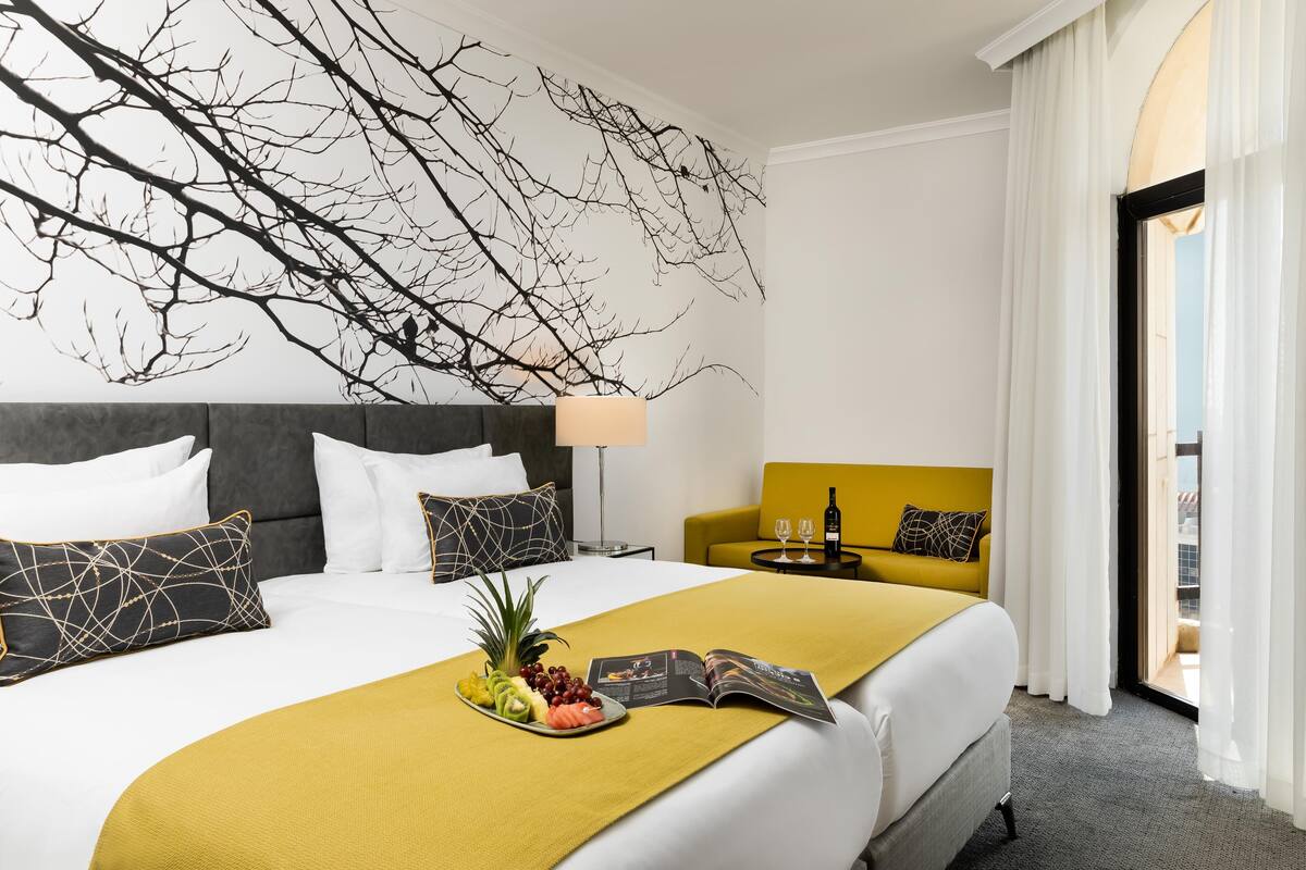 Quarto de hotel com uma cama grande, um sofá pequeno, uma mesa de jantar com uma garrafa e copos, e um prato de frutas sobre a cama.