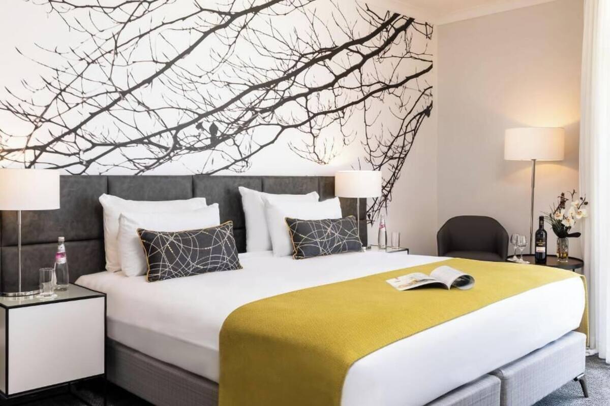 Quarto de hotel com uma cama grande, duas mesas de cabeceira, uma luminária, uma cadeira e uma obra de arte na parede.