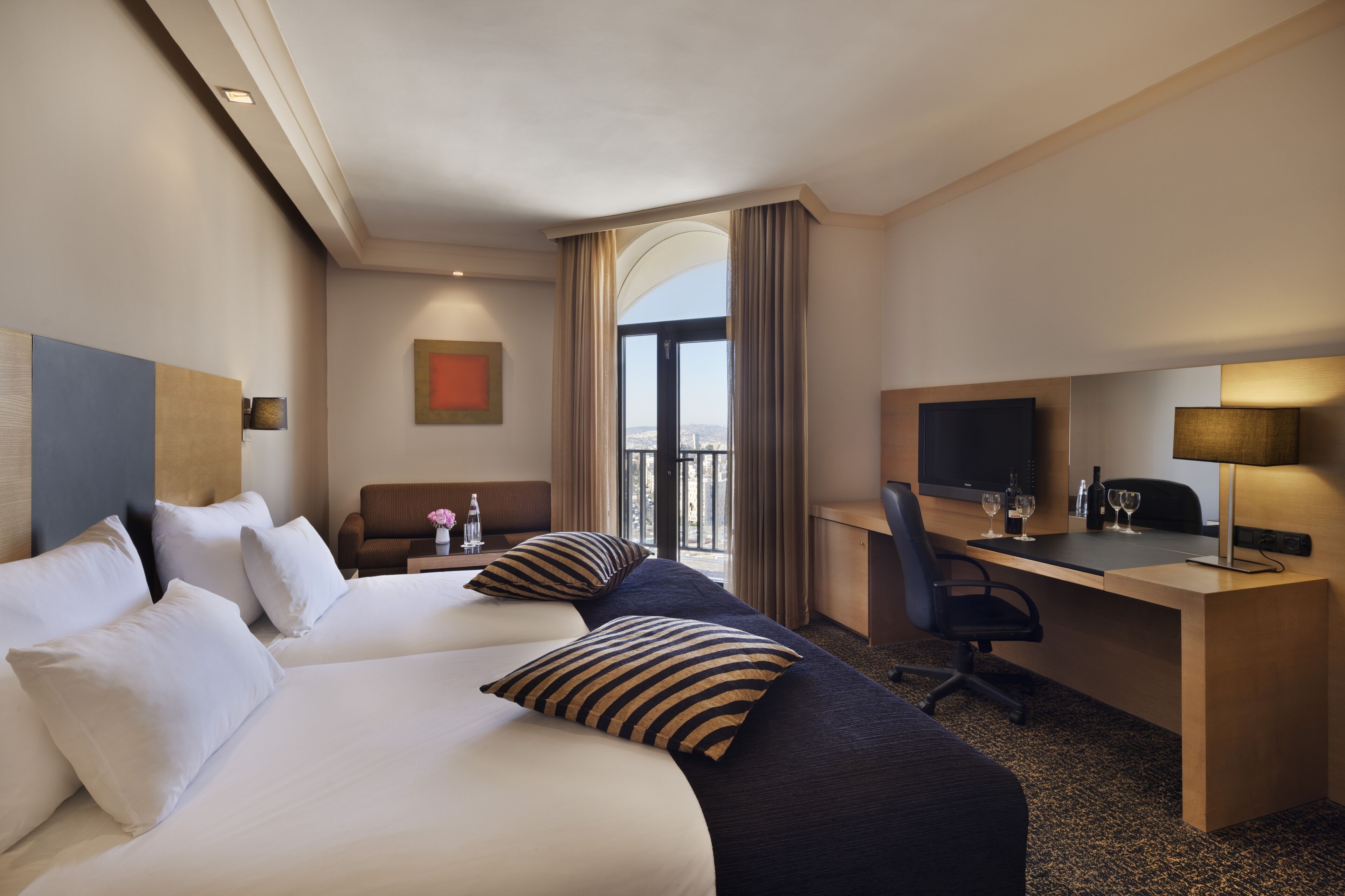 Quarto de hotel com duas camas, uma escrivaninha, uma cadeira, uma televisão e uma varanda com vista.