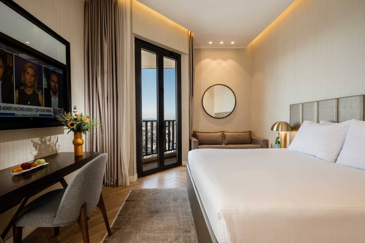 Quarto de hotel com uma cama grande, uma escrivaninha com cadeira, televisão na parede, espelho redondo e varanda com cortinas.
