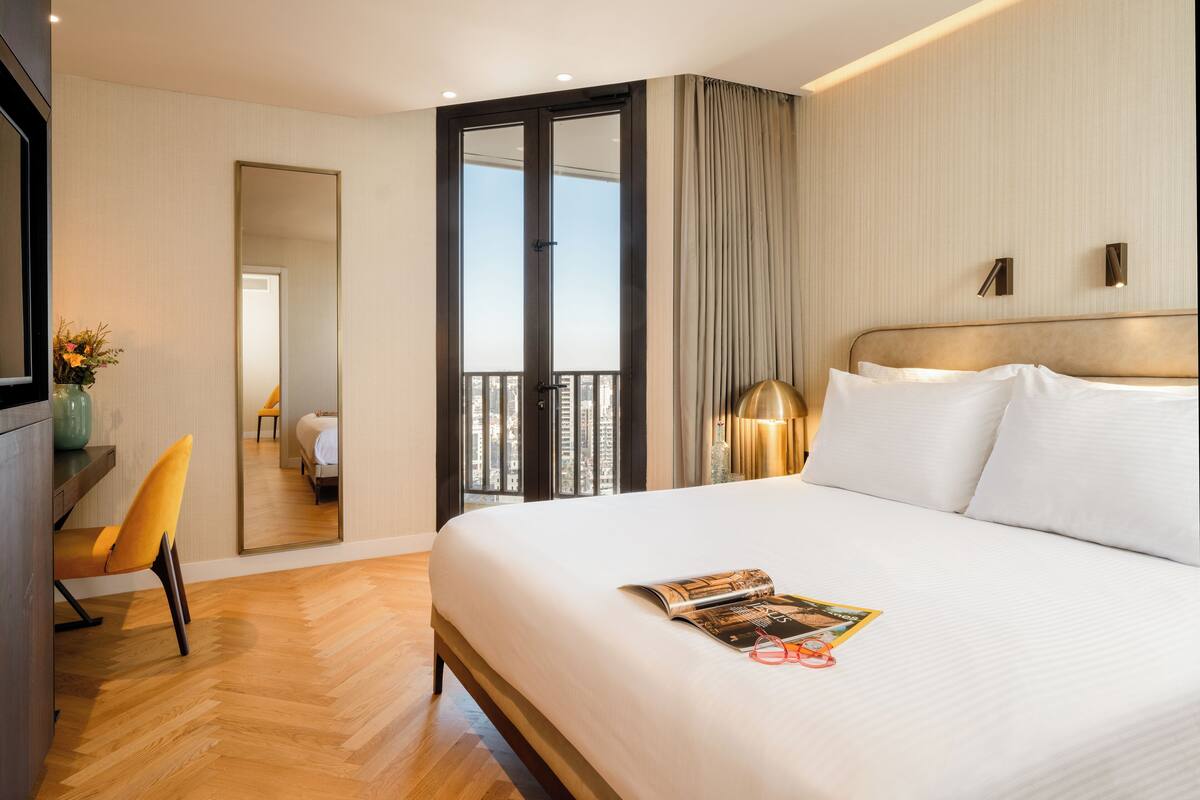 Quarto de hotel com uma cama grande, uma escrivaninha com cadeira, um espelho e vista para a cidade pela janela.