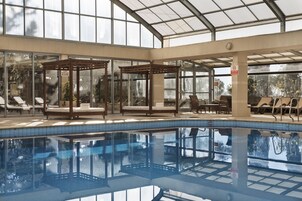 Piscina interior com paredes de vidro transparente, espreguiçadeiras e gazebos de madeira.