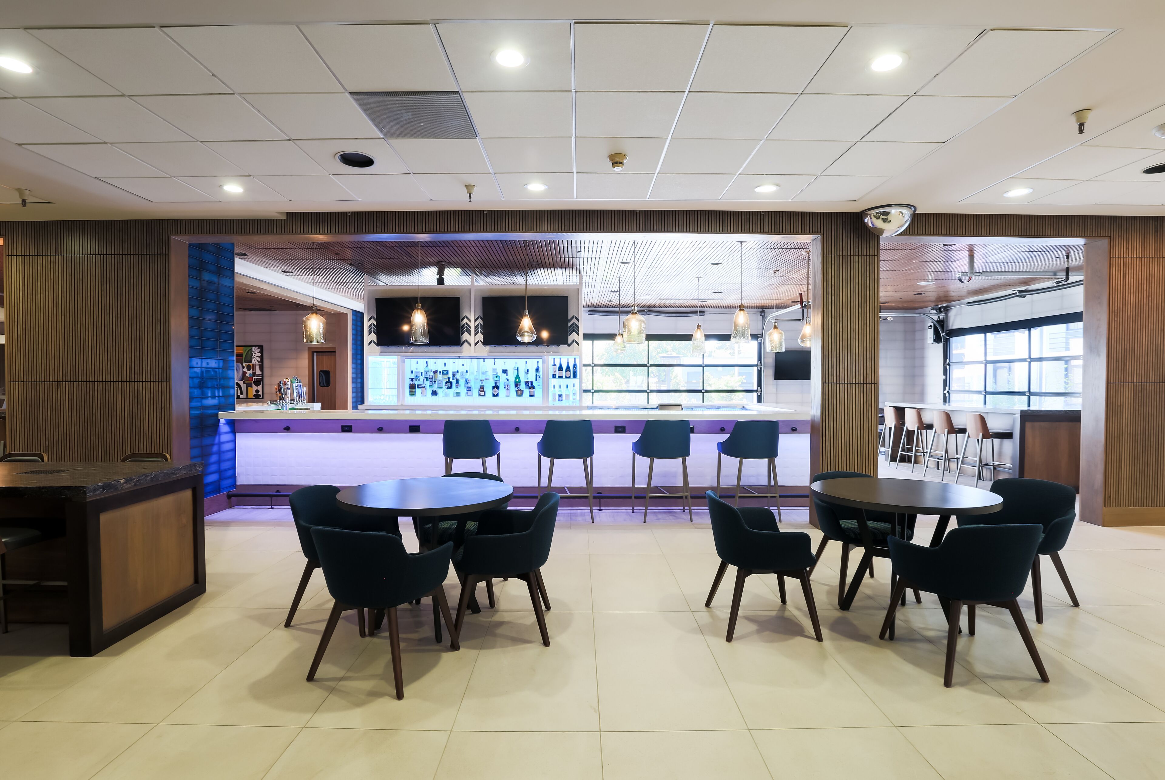 Restaurante com mesas e cadeiras redondas, área de bar com garrafas e design de teto moderno.