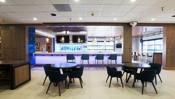 Restaurante com mesas e cadeiras redondas, área de bar com garrafas e design de teto moderno.