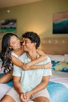 Um casal sentado na cama de um quarto de hotel, com uma parede lisa e um quadro ao fundo.