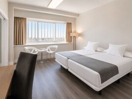Quarto de hotel com cama, escrivaninha, duas cadeiras, abajur e janela com cortinas.