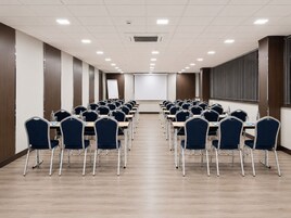 Uma sala de conferências com mesas e cadeiras dispostas em fileiras, uma lousa e uma tela de projeção.