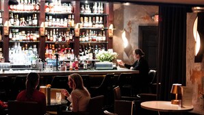Uma área de bar com uma placa de neon que diz "The Irving", prateleiras de madeira repletas de garrafas e clientes sentados em mesas.