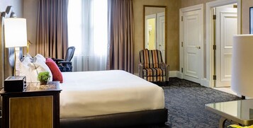 Quarto de hotel com uma cama, uma escrivaninha com luminária, uma cadeira e uma porta.