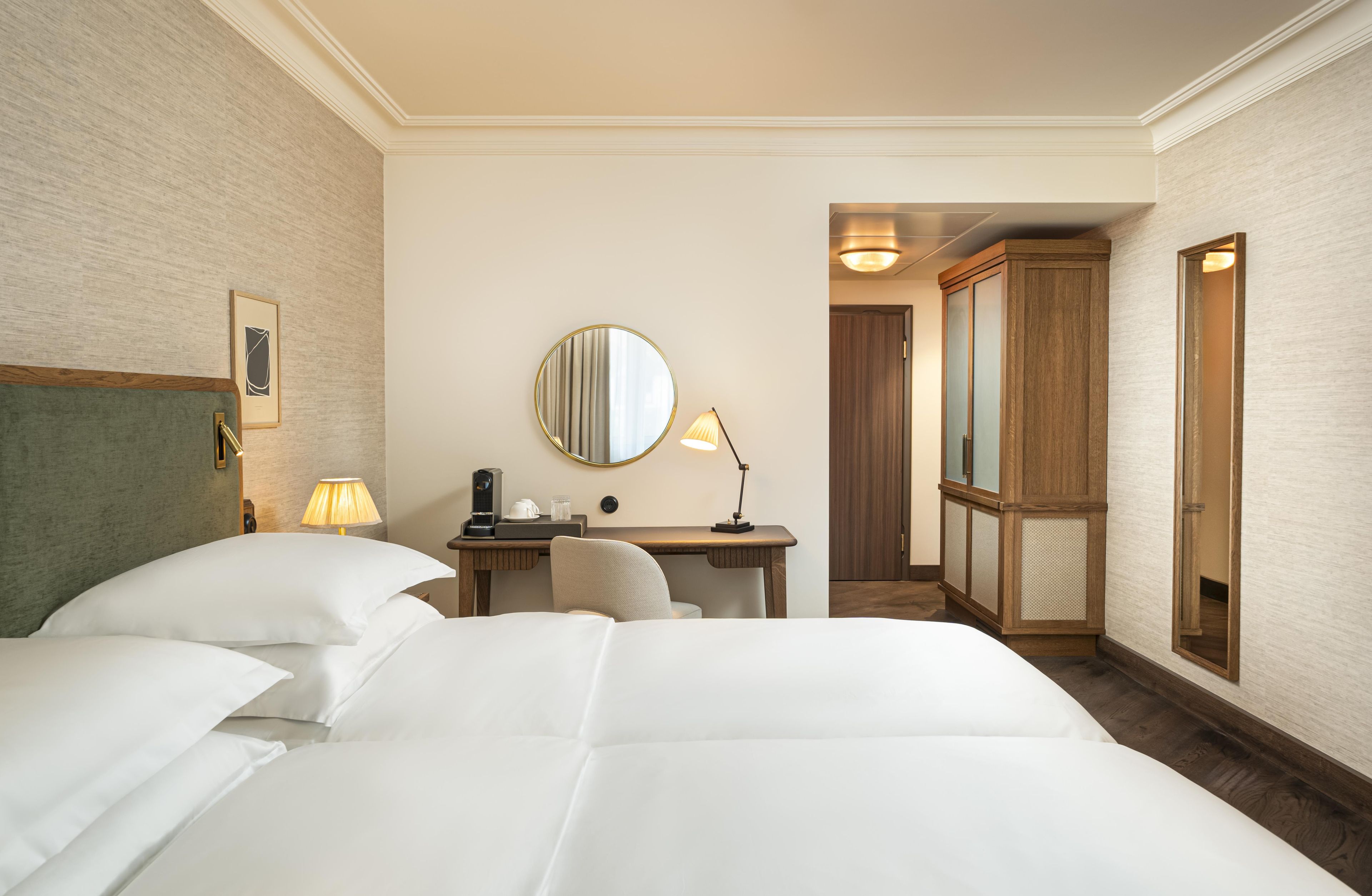 Quarto de hotel com uma cama grande, uma escrivaninha, uma cadeira, um espelho redondo, um abajur e um guarda-roupa.