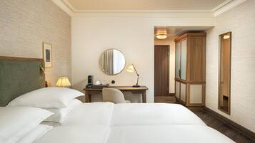 Quarto de hotel com uma cama grande, uma escrivaninha, uma cadeira, um espelho redondo, um abajur e um guarda-roupa.